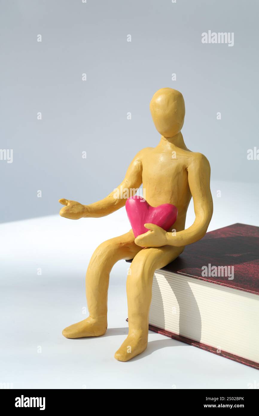 Yellow plasticine human figure with pink heart sitting on book against white background Stock ...