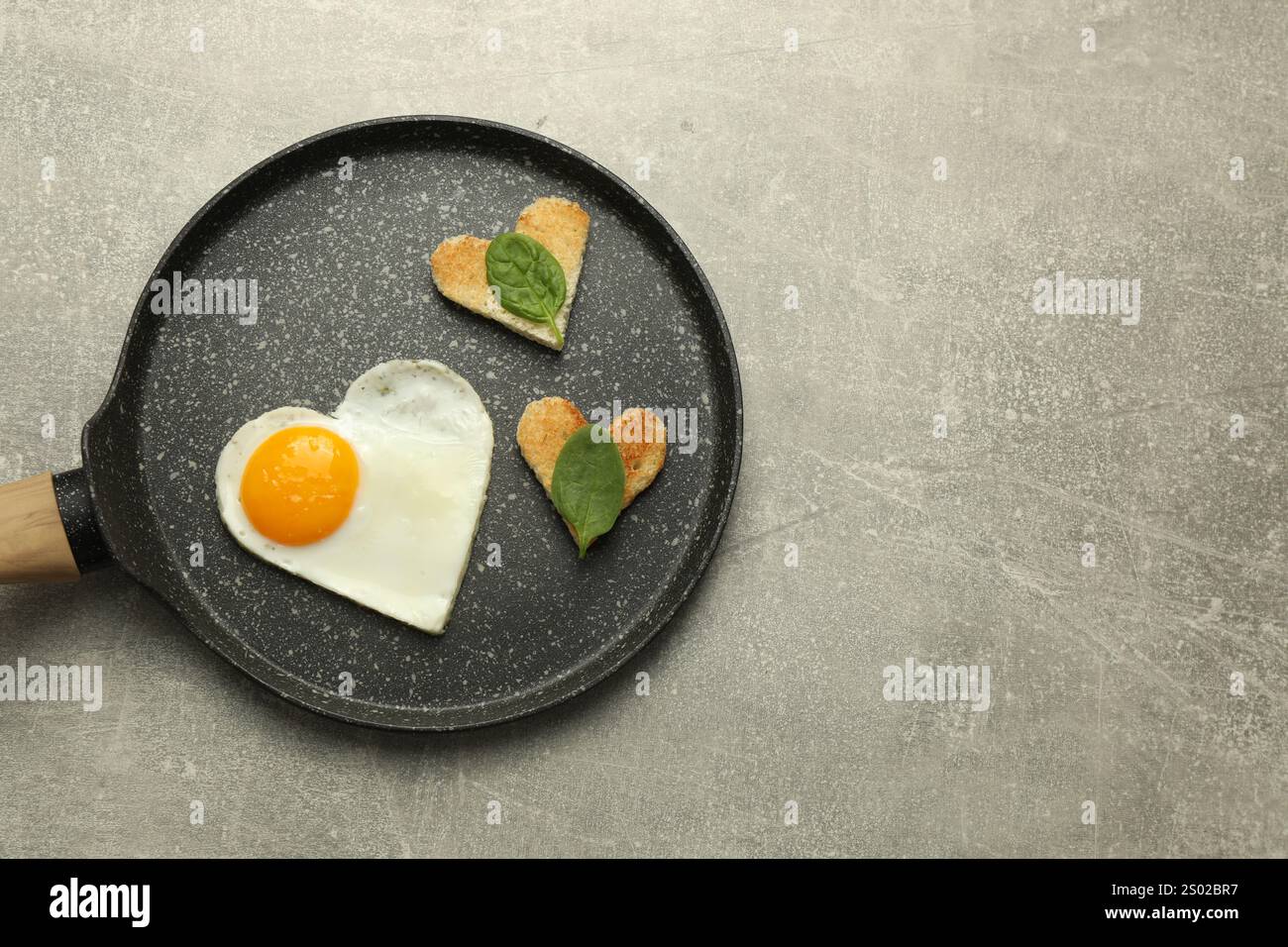 Heart shaped fried egg and toasts in frying pan on grey marble table, top view. Space for text ...