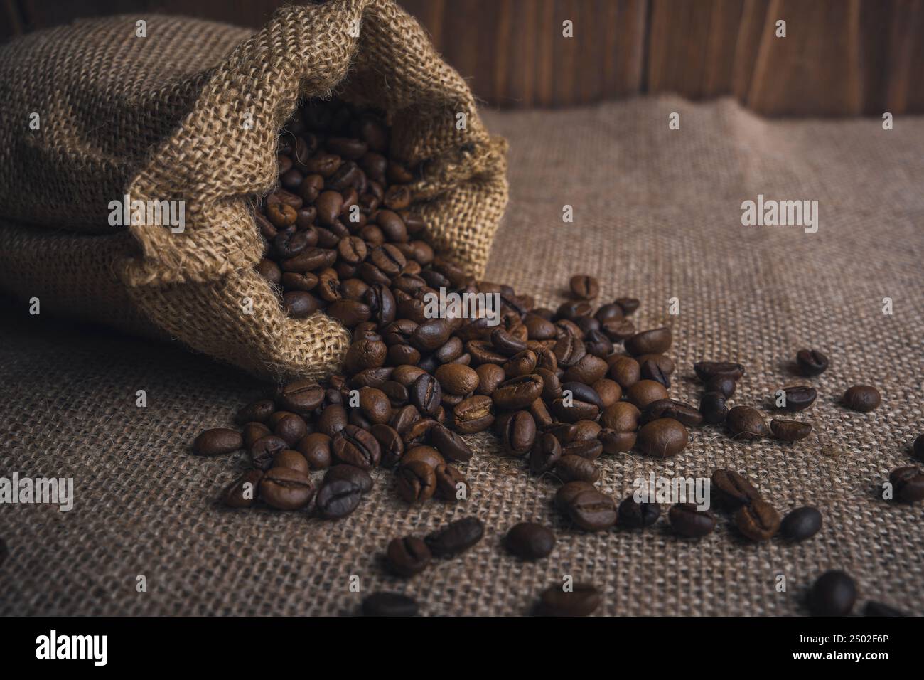 A burlap sack spilling roasted coffee beans onto a rustic burlap fabric surface with a wooden ...