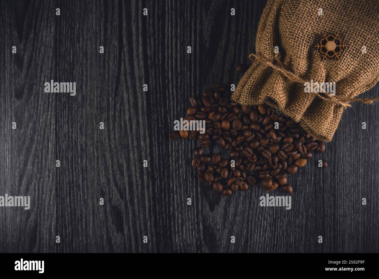 A burlap sack spilling roasted coffee beans onto a textured dark wooden surface with a rustic ...