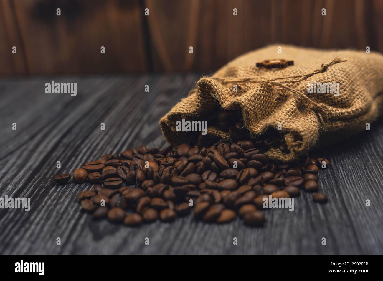 A rustic burlap sack spilling roasted coffee beans onto a textured dark wooden surface, creating ...