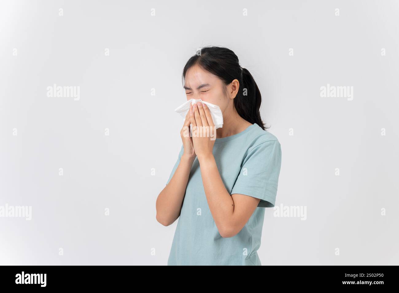 A sick, uncomfortable young Asian woman sneezing and blowing her nose ...
