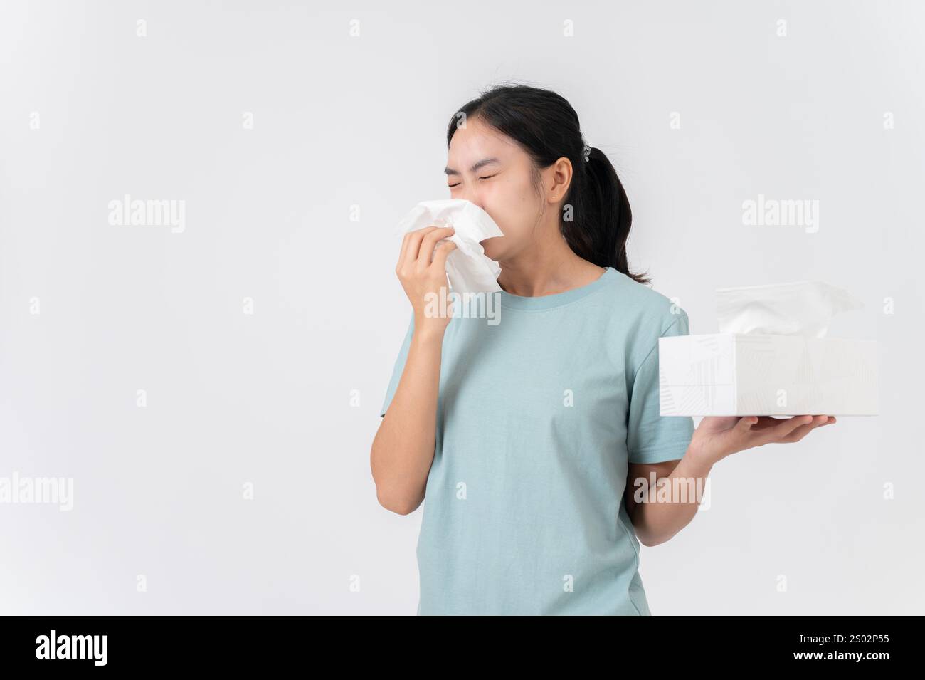 A sick, uncomfortable young Asian woman holding a tissue box, sneezing ...