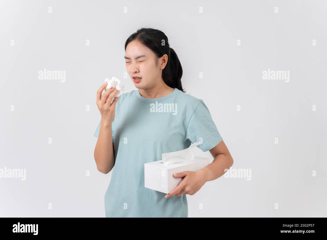 A sick, uncomfortable young Asian woman holding a tissue box, sneezing ...
