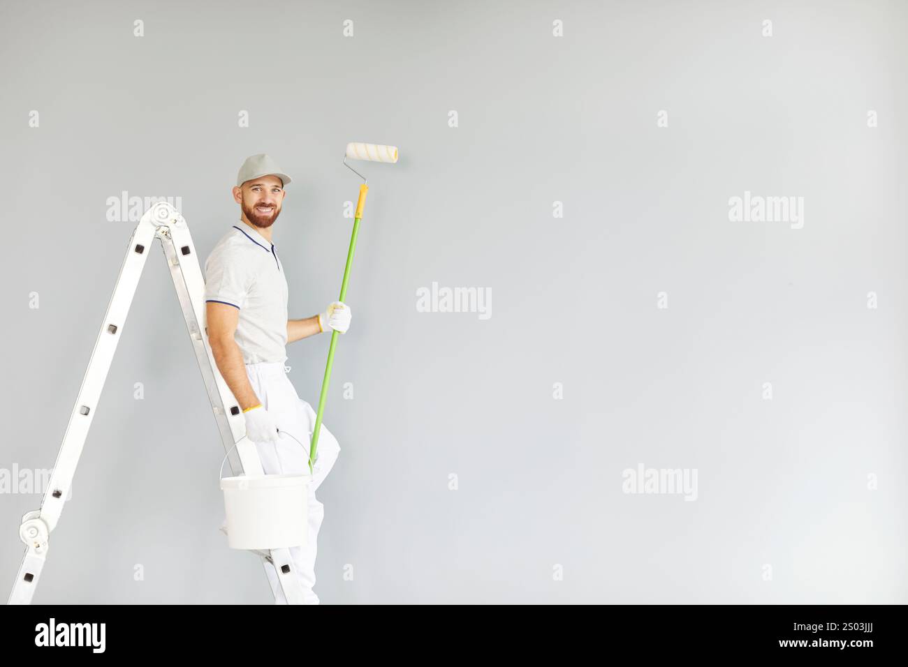 Smiling painter in white attire standing on ladder with roller and ...