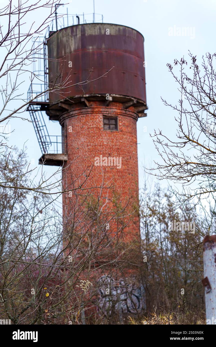 An old red brick water tower with a rusted metal tank and overgrown ...