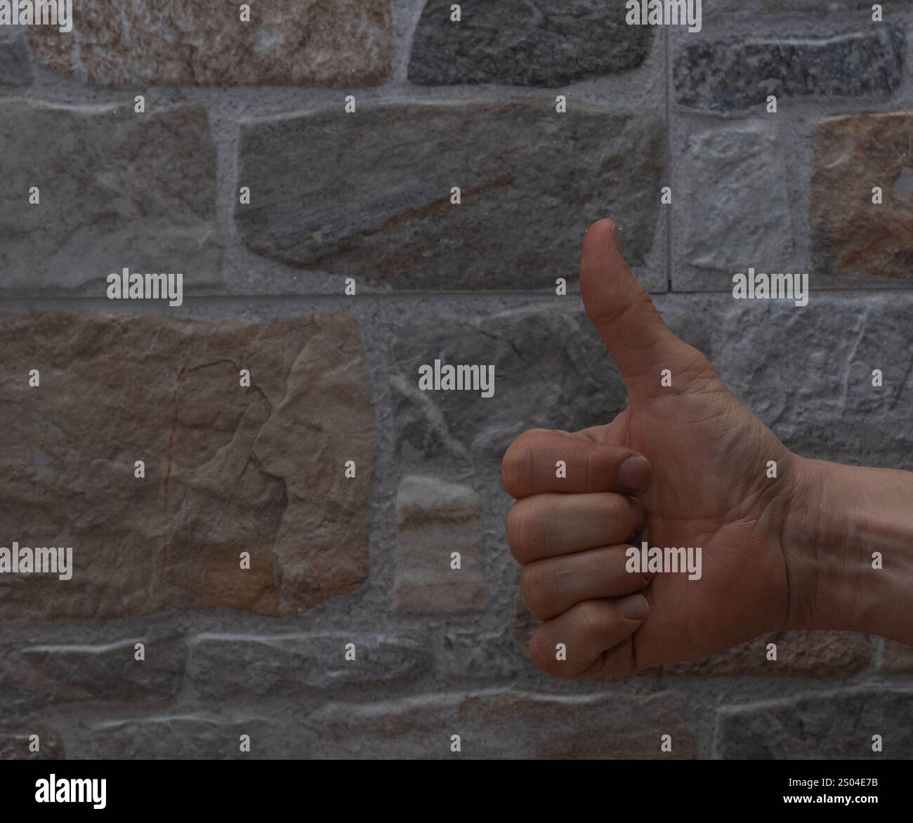 A human hand displays a thumbs up sign against a textured stone wall ...
