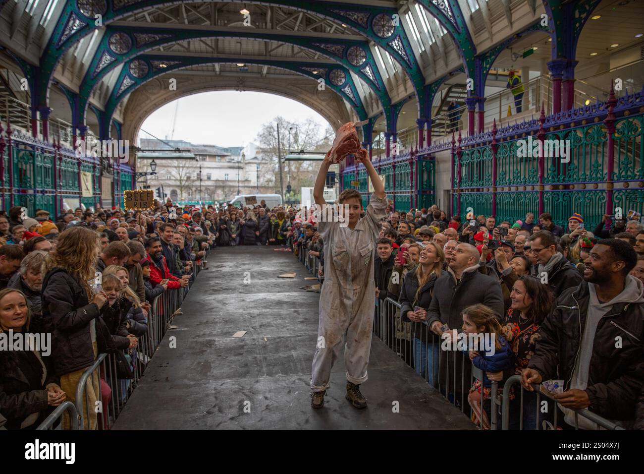 A young butcher holds up a joint of meat in-front of a crowd. In a ...