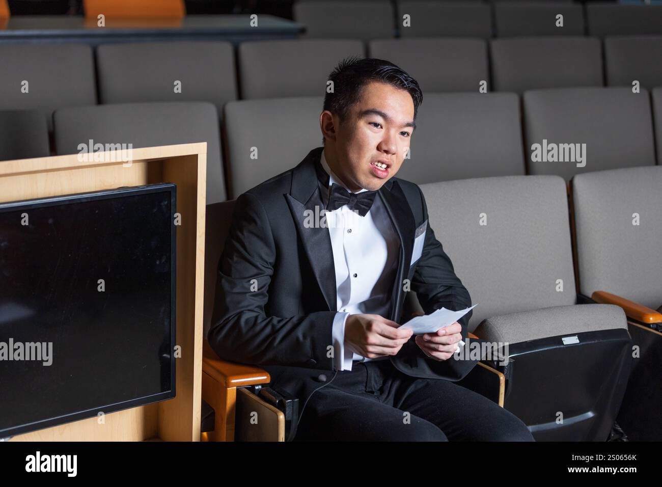 A young university student is at a public speaking competition, seated in a auditorium hall ...