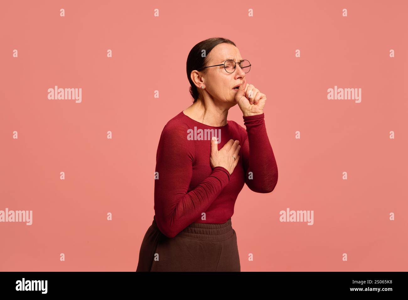 Woman in burgundy top coughing into fist, other hand on chest, eyes ...