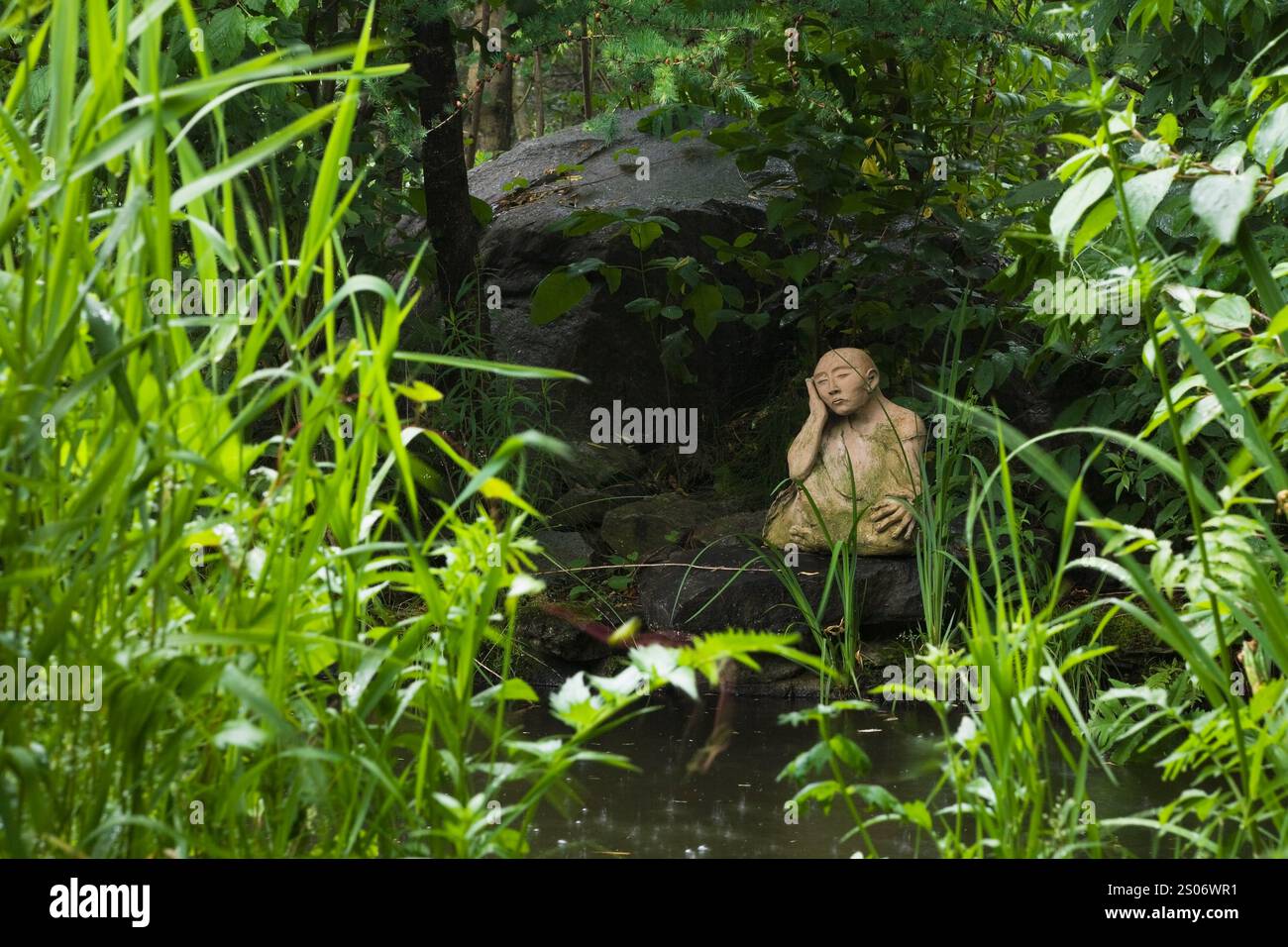 Ornamental beige Buddha statue through green plant leaves next to pond ...