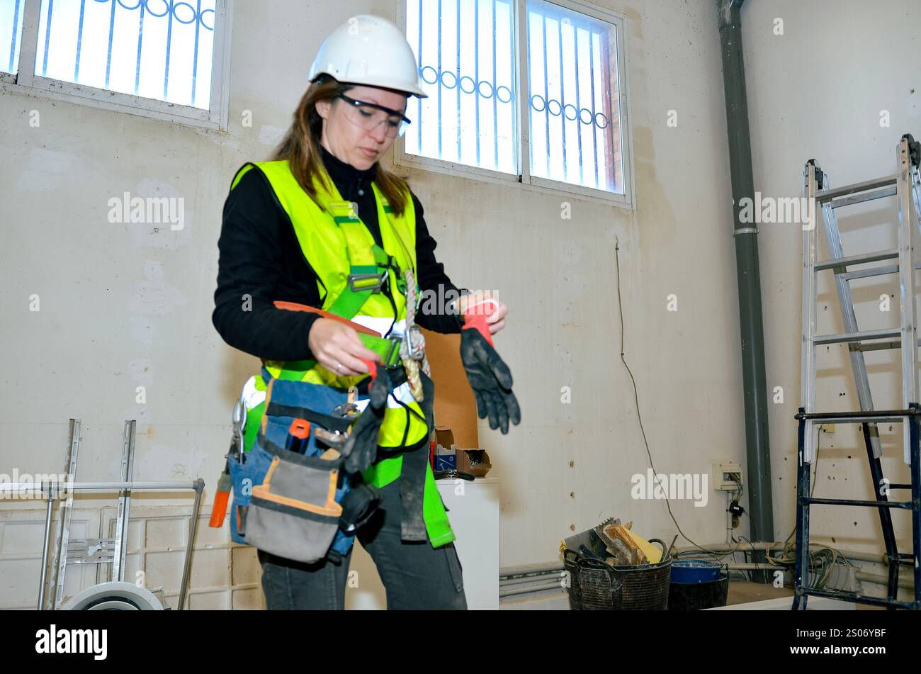 Female construction worker wearing safety helmet and vest putting on gloves at a construction ...