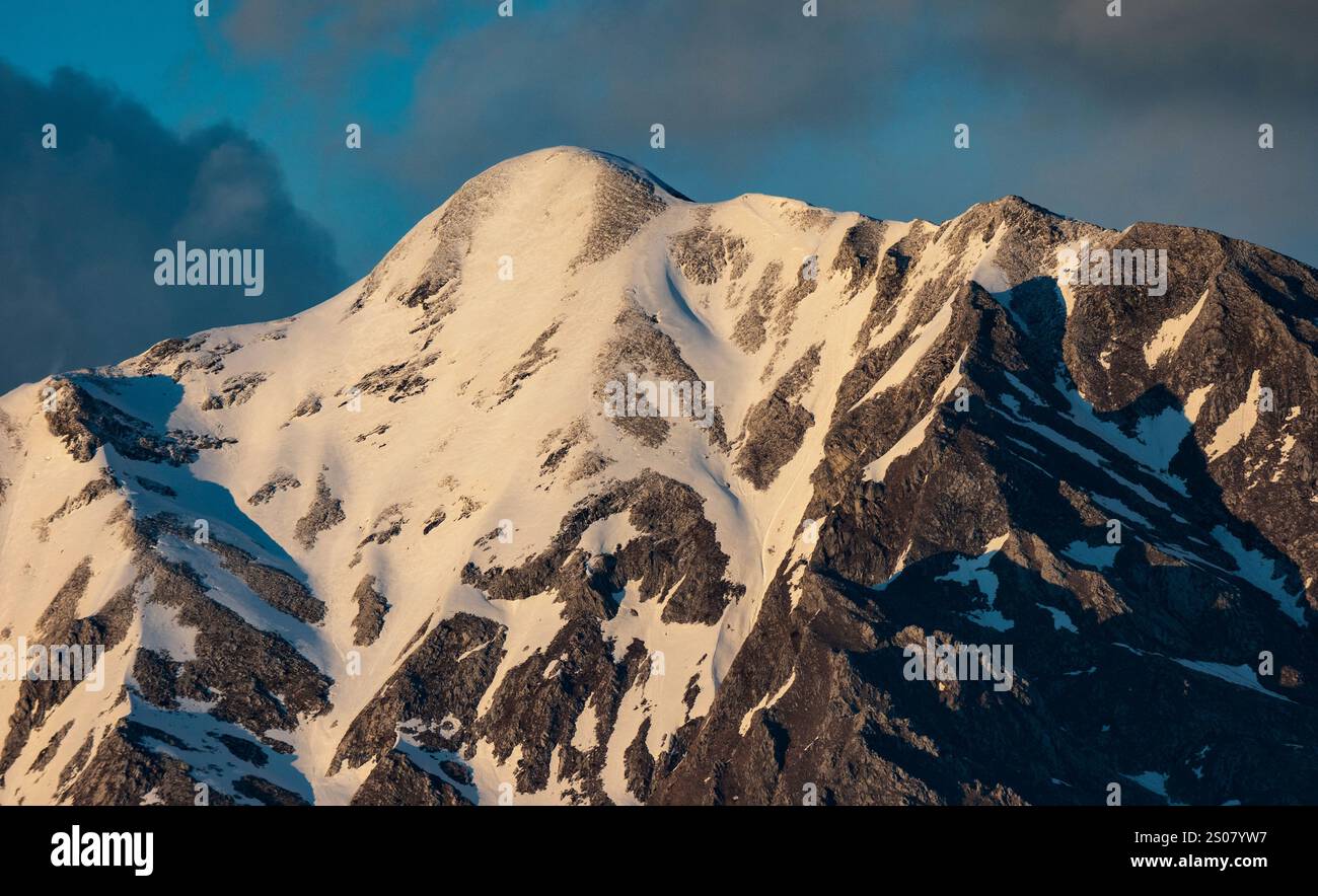 A majestic snow-capped mountain peak under a blue sky with clouds. The sunlight highlights the ...