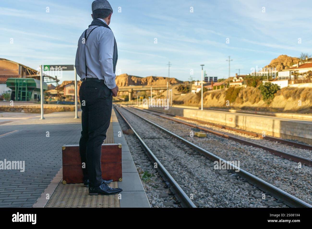 A man alone on the platform next to the train tracks, his suitcase on ...