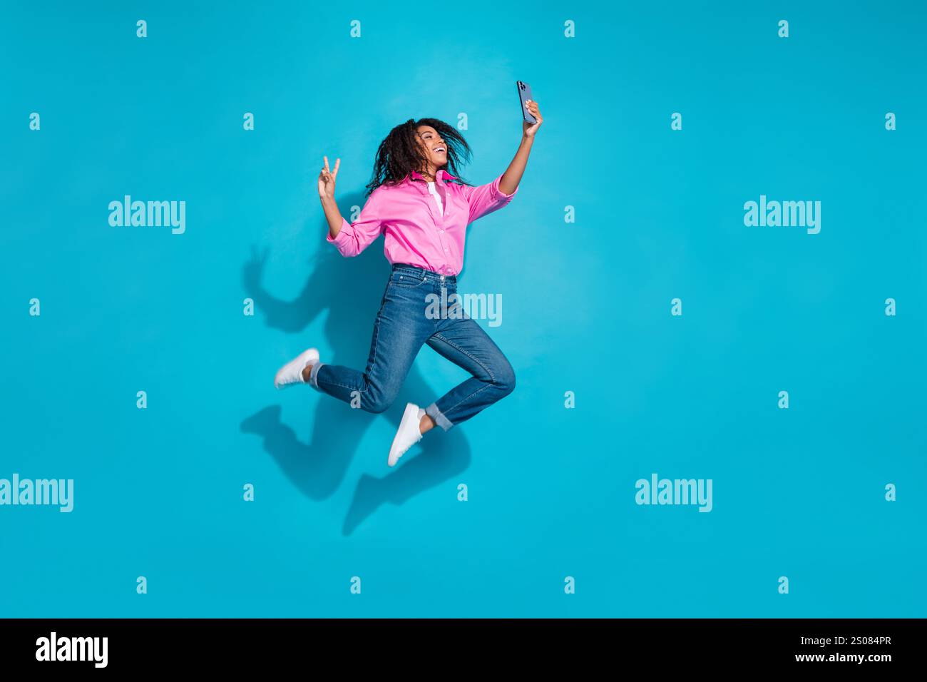 Joyful young woman jumping with peace sign, holding smartphone against ...