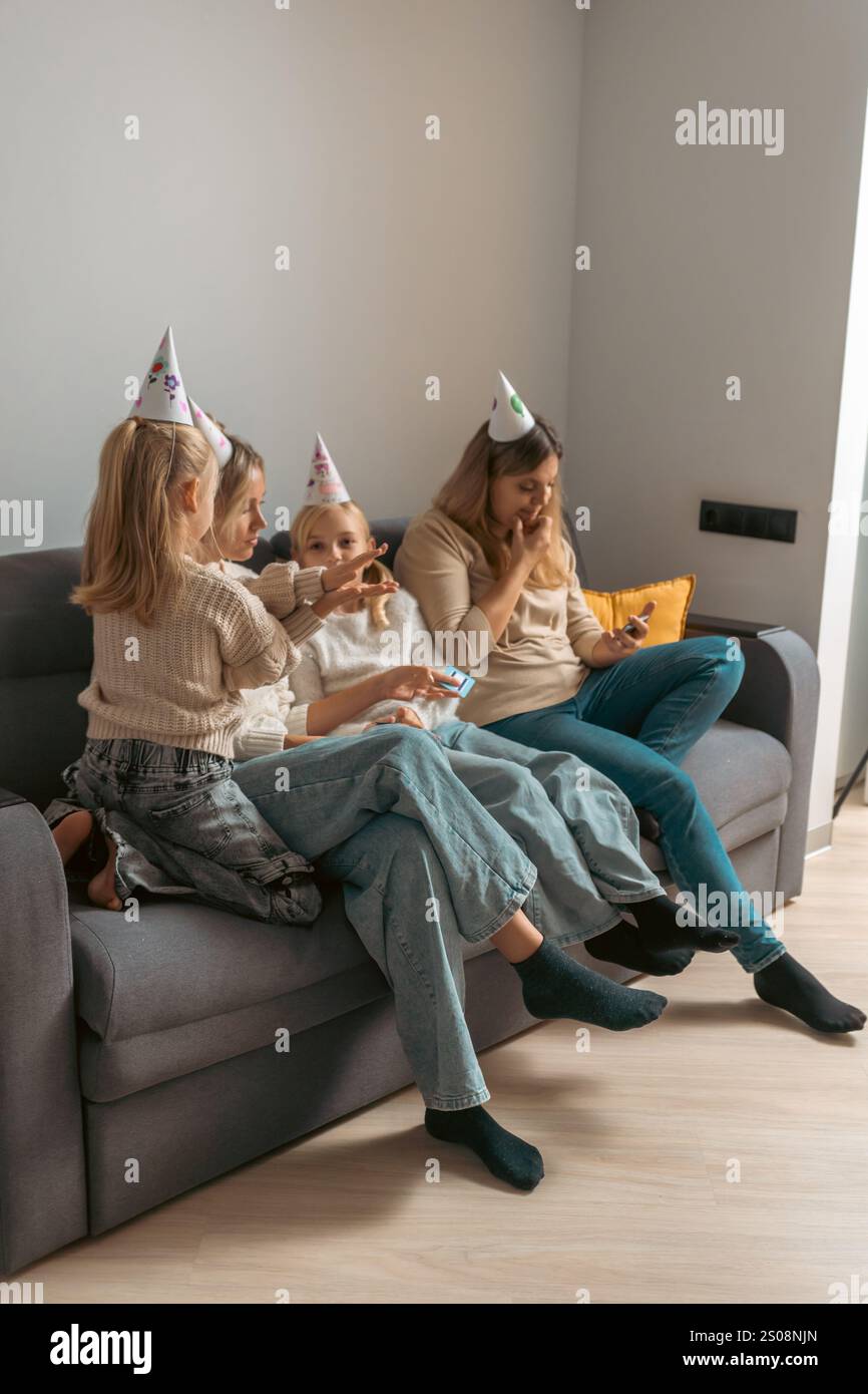 Four women, wearing party hats, sit on a gray couch, eating and ...