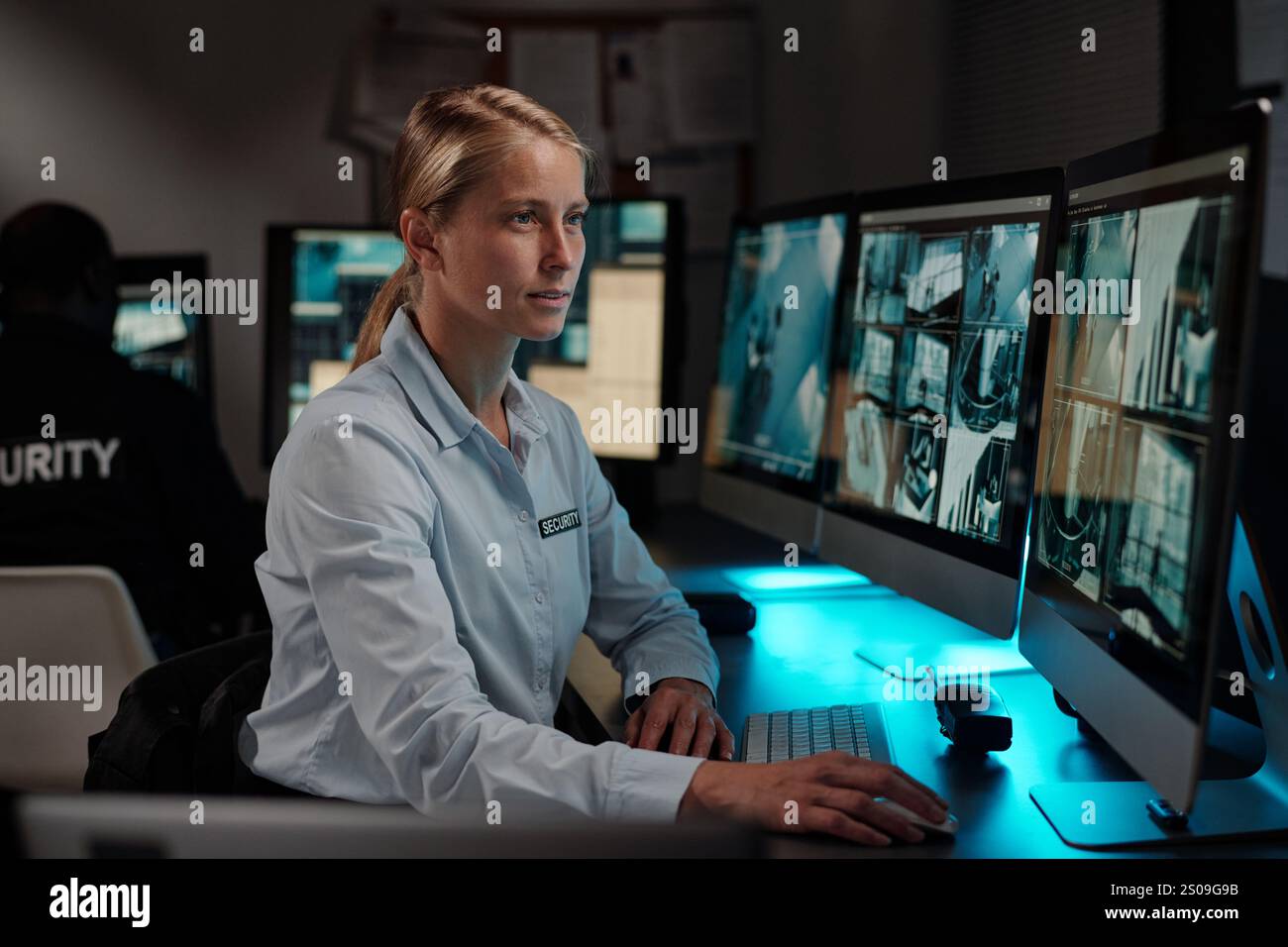 Young blond female security guard in uniform sitting by desk in control ...