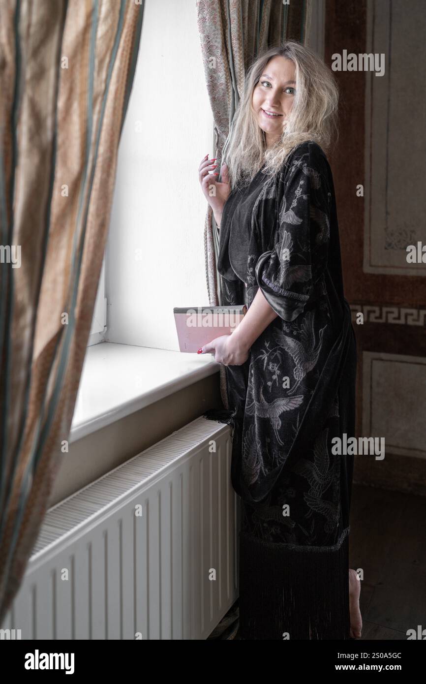 A woman standing near a window, clad in a dark patterned cloak, holding ...