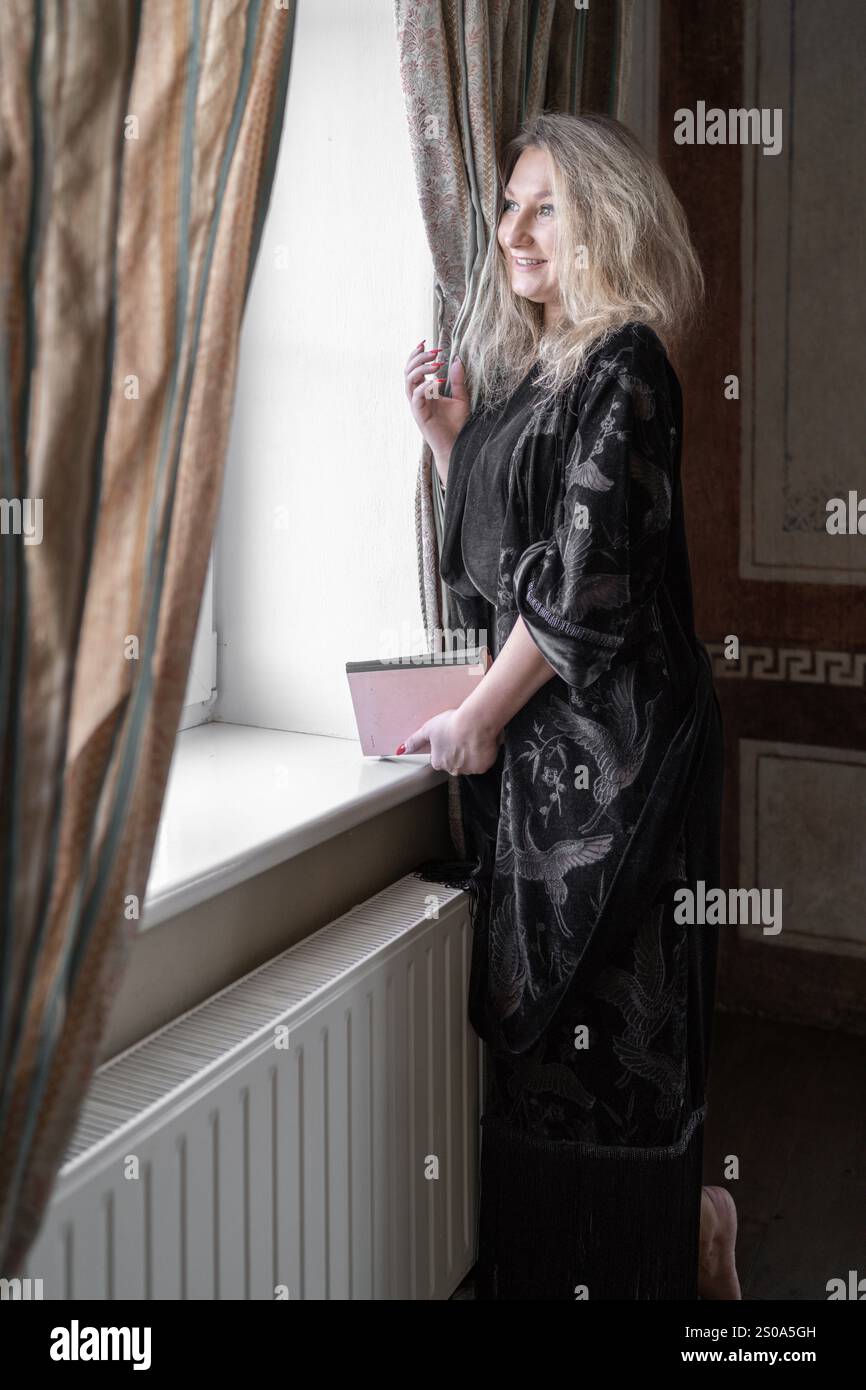 A woman standing near a window, clad in a dark patterned cloak, holding ...