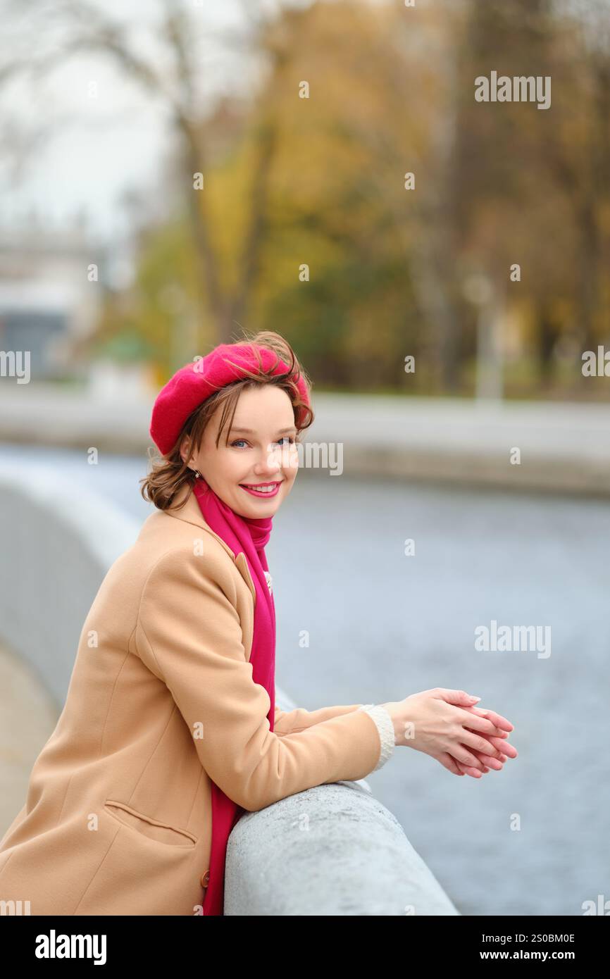 A joyful woman wears a red beret and beige coat while resting her hands ...