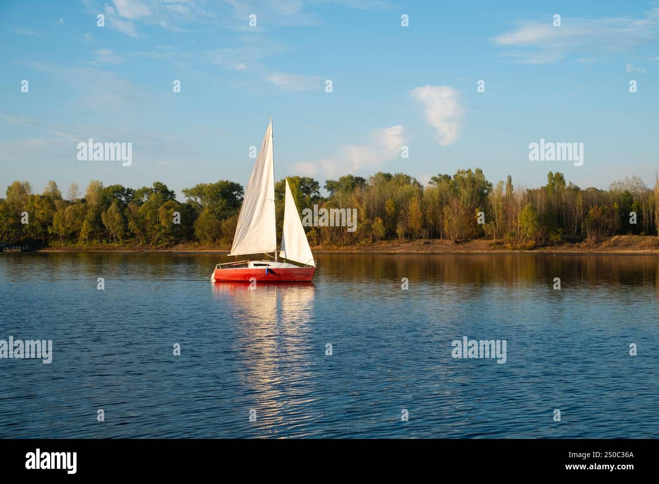 A small red boat is sailing on a lake. The boat is white and has a sail ...