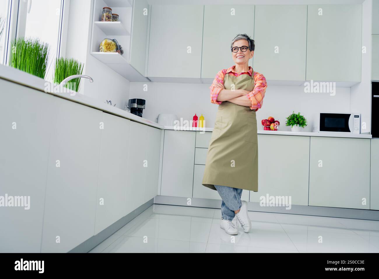 A cheerful elderly woman stands confidently in a bright kitchen ...