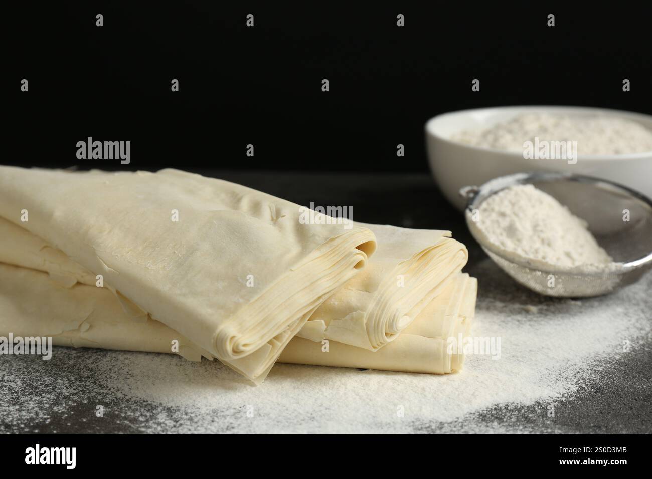 Raw puff pastry dough and flour on dark table against black background ...
