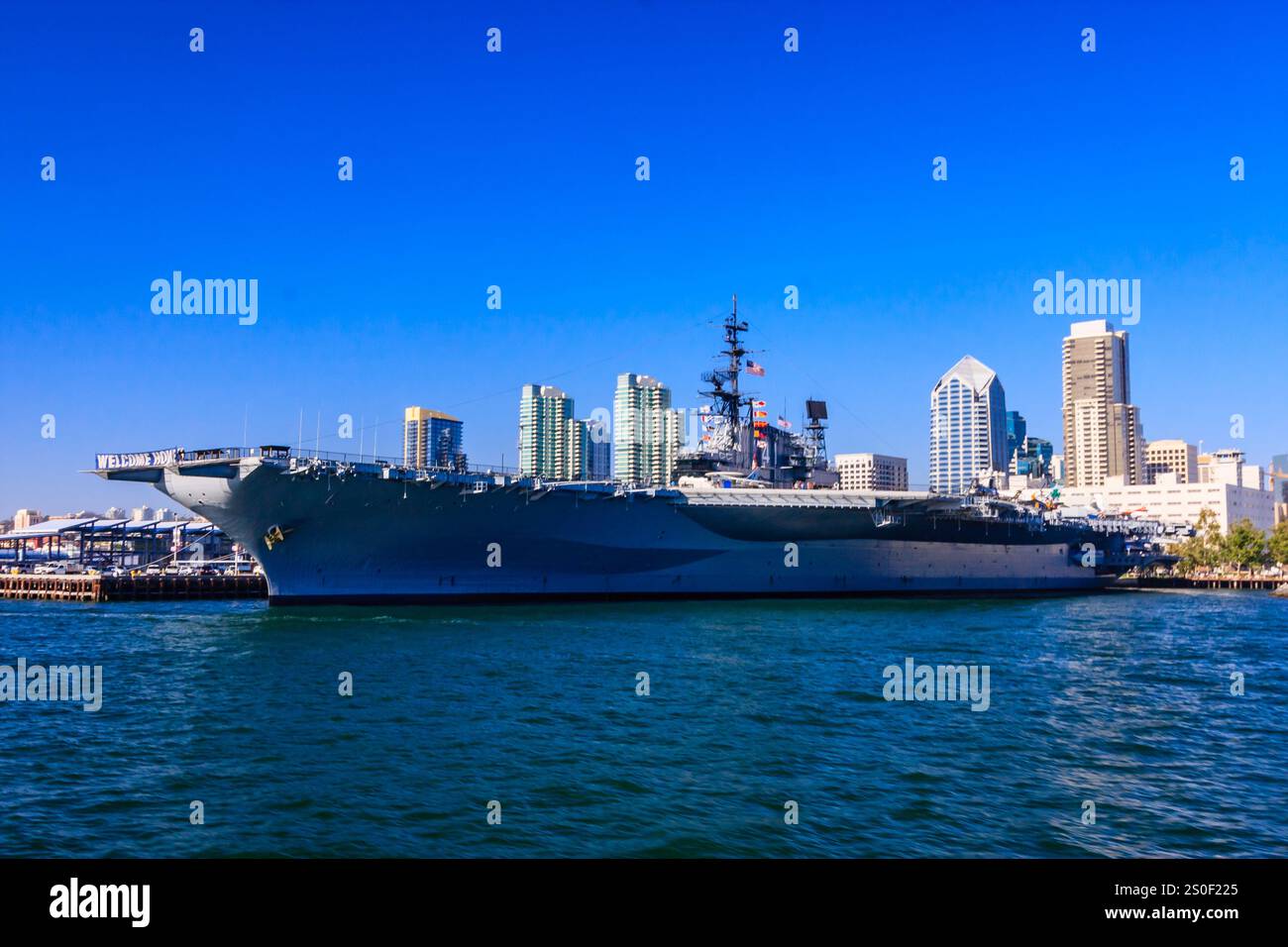 A large navy ship is docked in a harbor next to a city. The sky is ...
