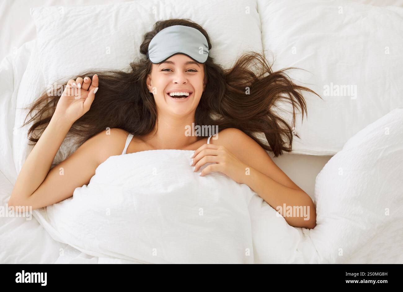 Well rested young woman wearing eye mask, lying in bed, waking up early ...