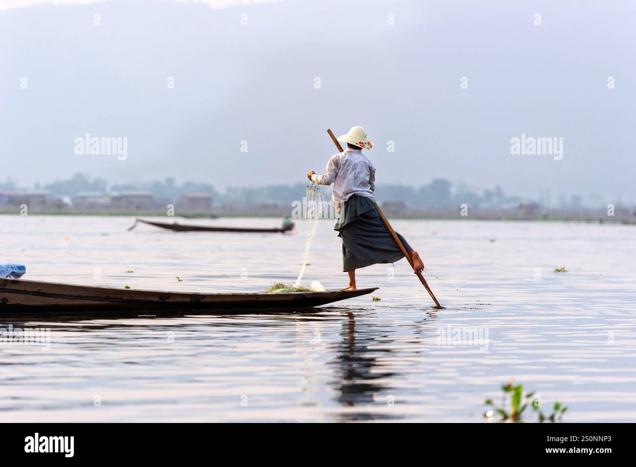 A woman standing on a wooden boat, casting a fishing net in a calm lake ...