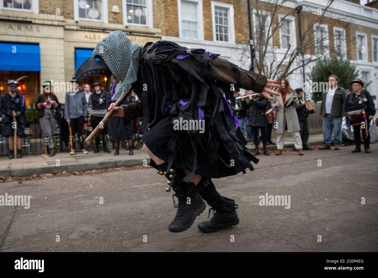 A Morris Dancer wearing a black mask runs along a street. Several ...