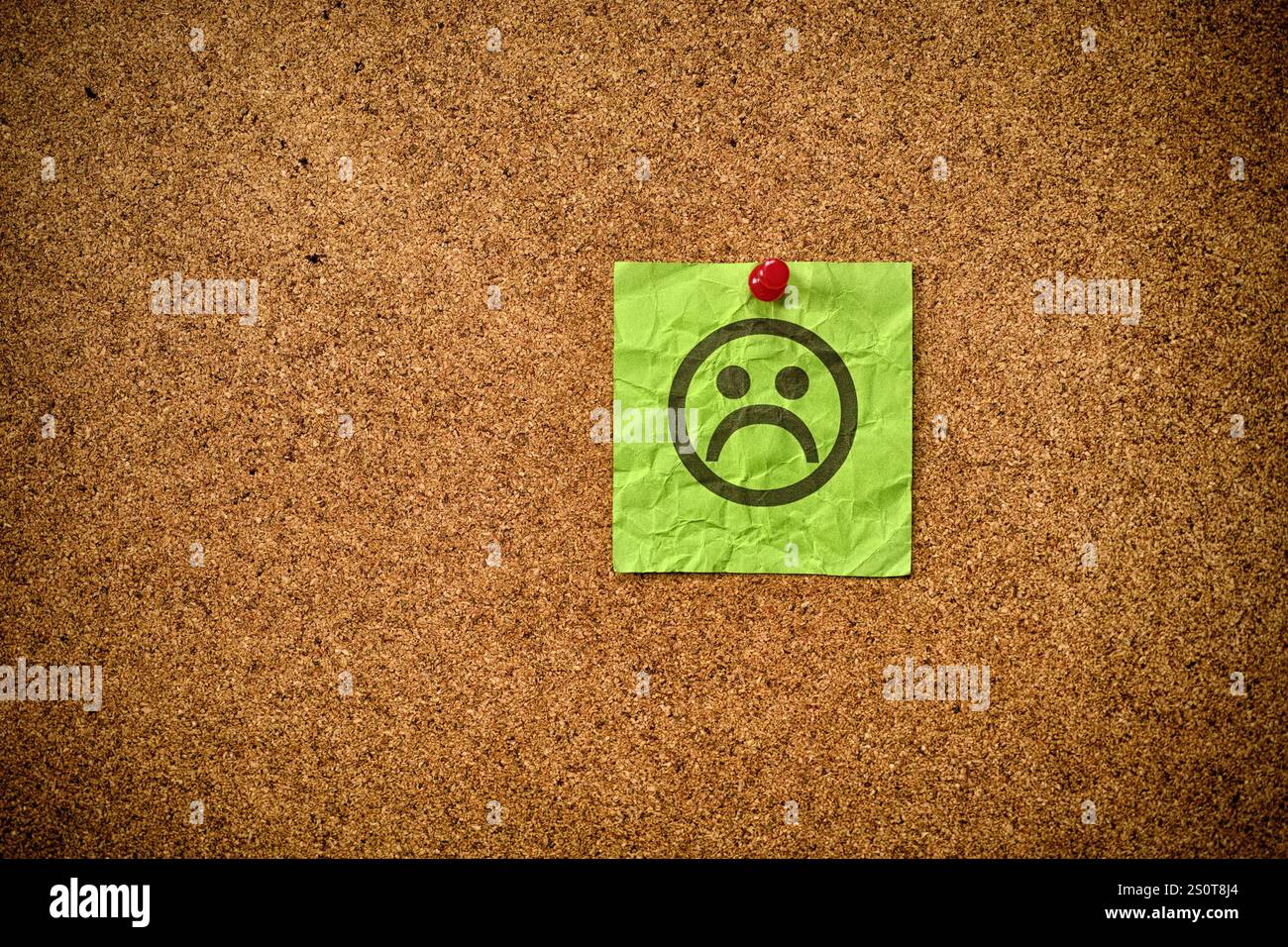 A green sticky note with a sad face on it pinned to a corkboard ...