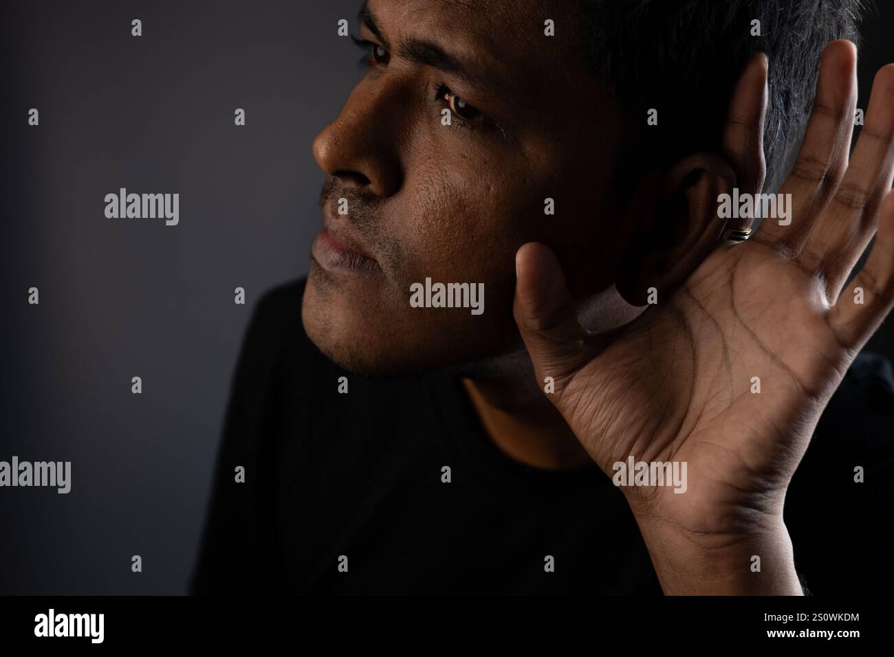 Man cups hand to ear, straining to hear a faint sound in the shadows. A portrait of focused ...