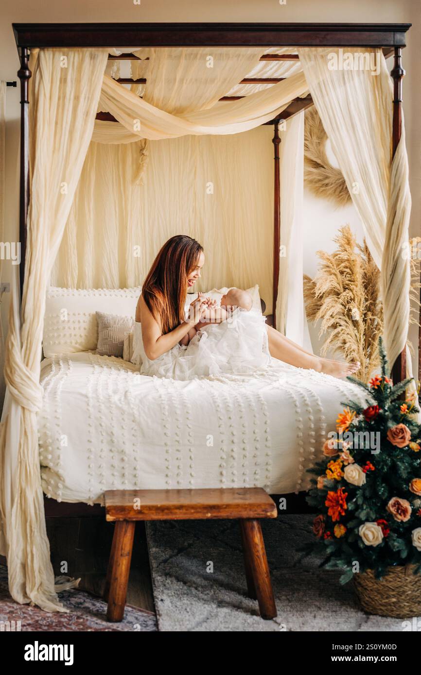 Mother in a white dress holding her baby on a cozy canopy bed Stock Photo - Alamy