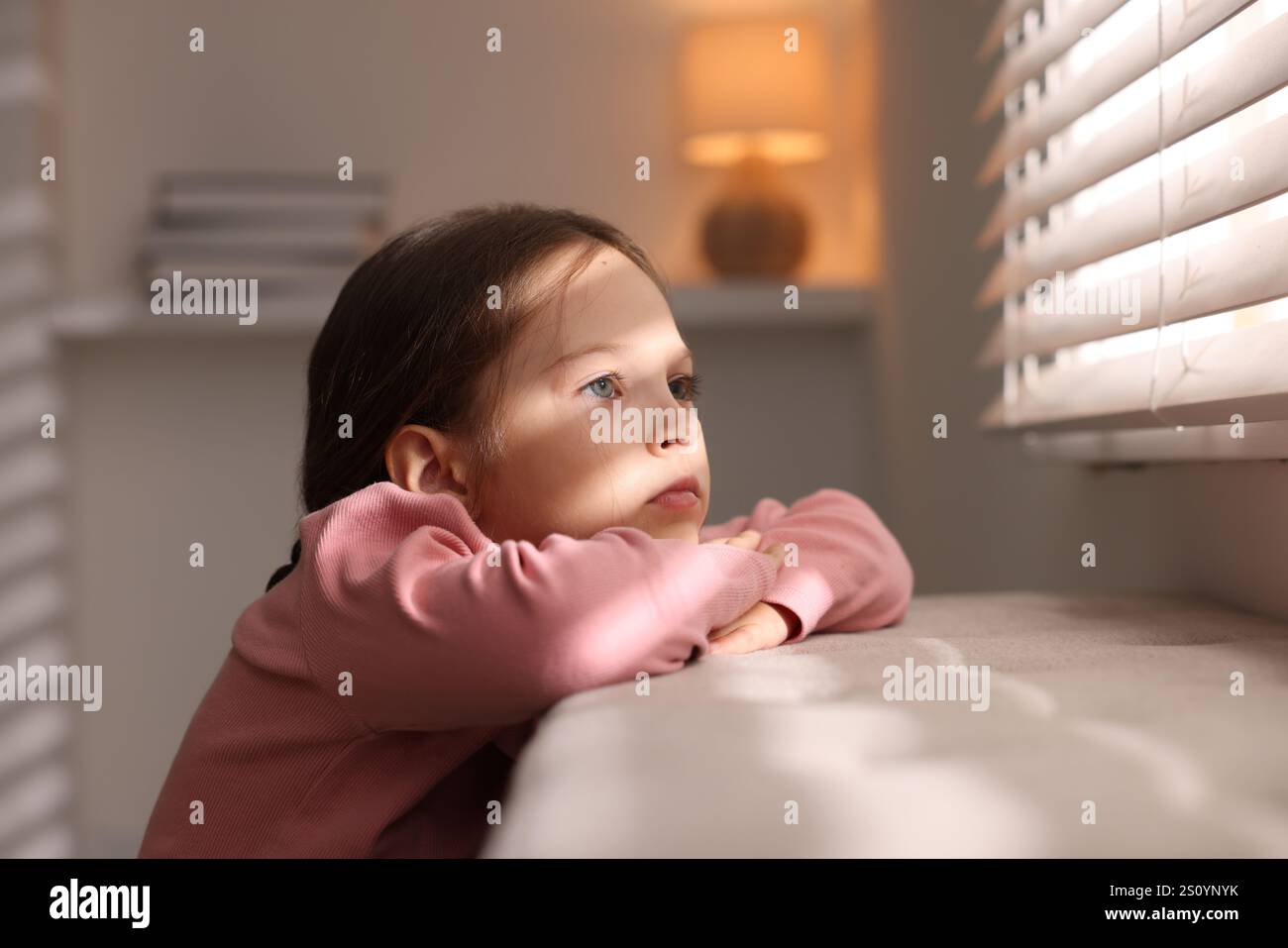 Orphanage concept. Sad girl and looking through window blinds indoors ...