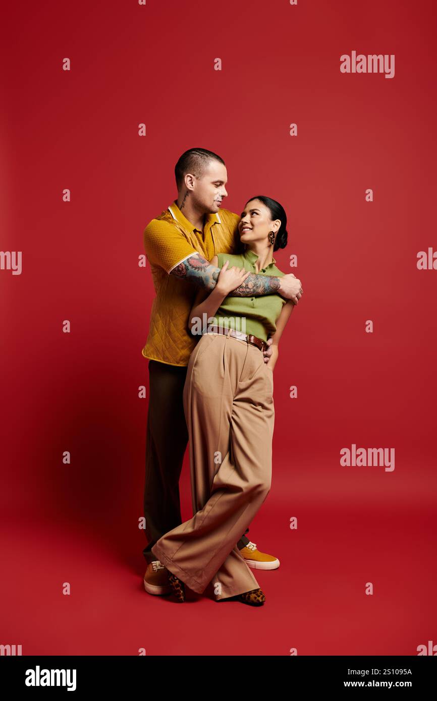 A joyful couple embraces in stylish outfits against a bold red studio ...