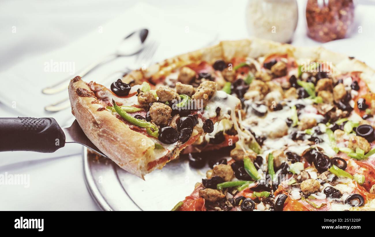 A view of a supreme style pizza pie, with a pie server lifting one of ...