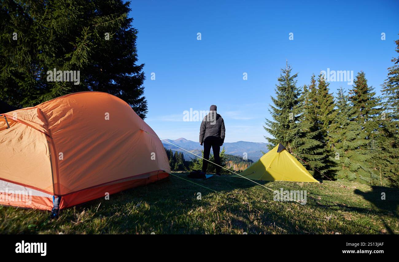 Back view of camper stands between orange and yellow tent, gazing at ...