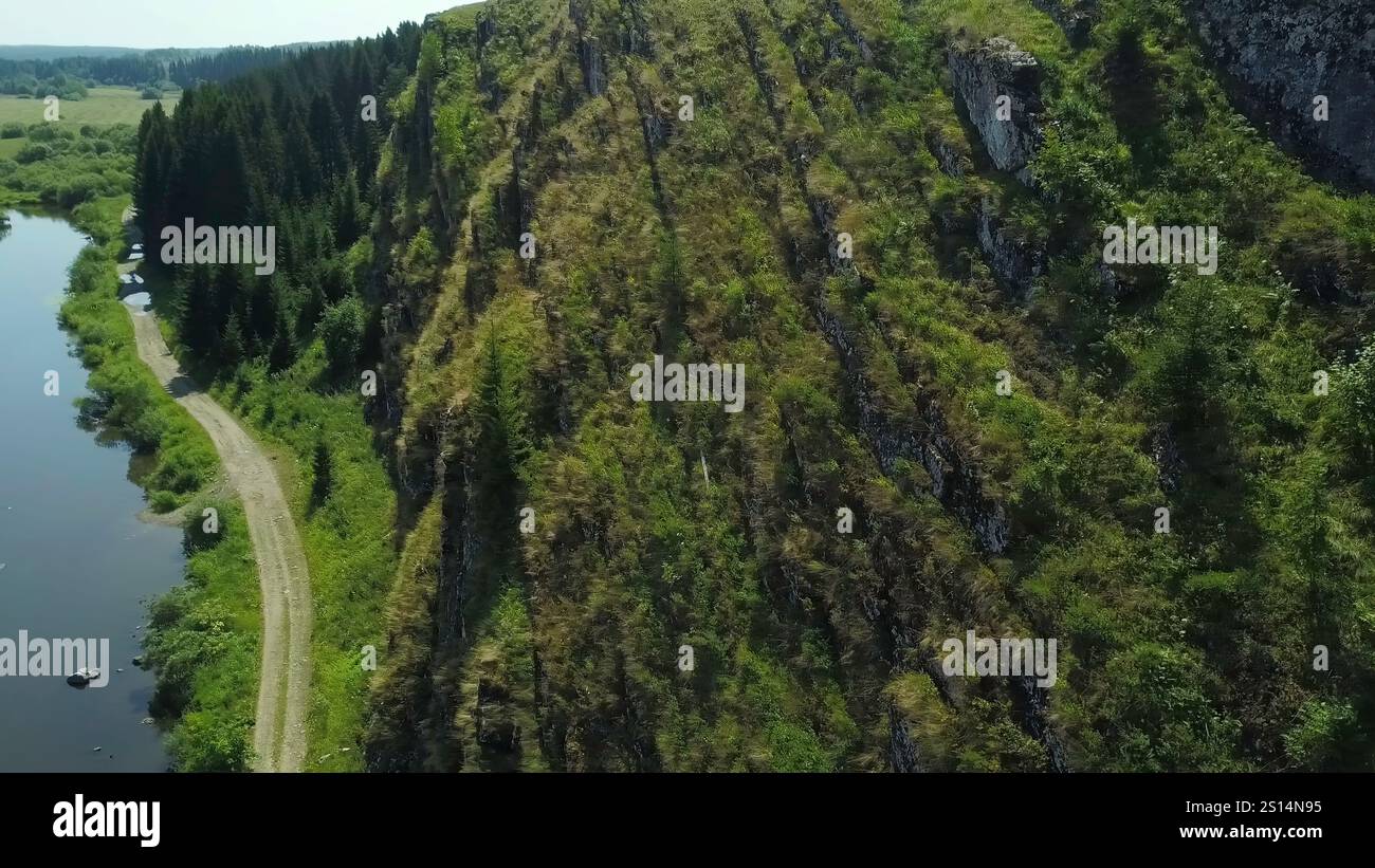 An aerial view shows a lush green cliff by a calm river, creating a ...
