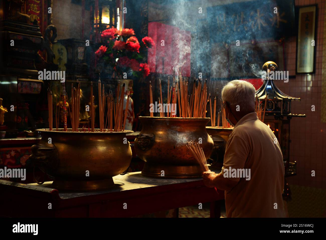An elderly man places incense sticks in a bronze urn inside a ...