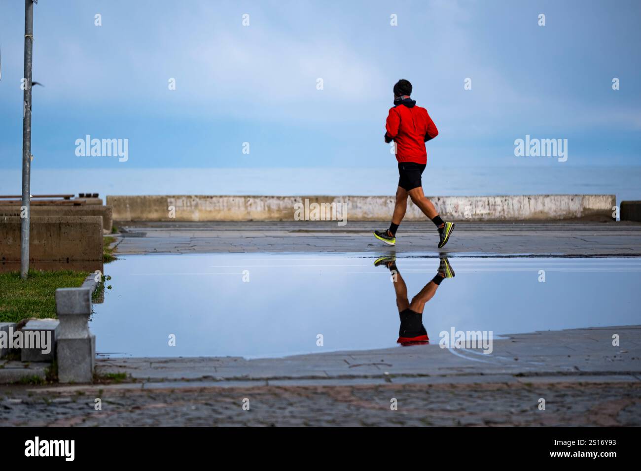A man in a red jacket is running on a wet sidewalk. The water is ...