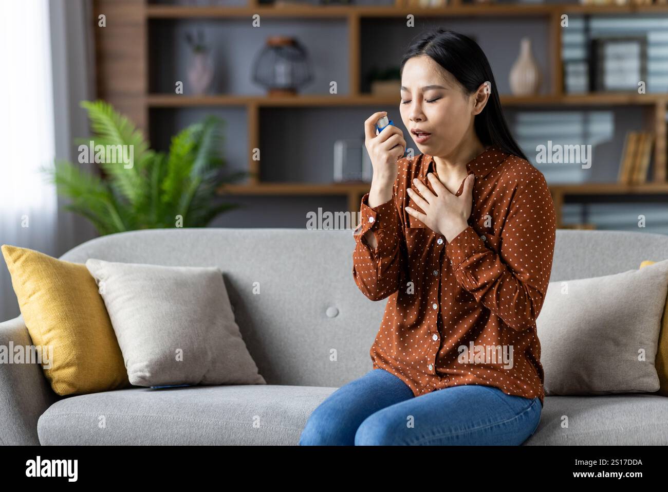 Asian woman using asthma inhaler, sitting on sofa experiencing breathing issue. Wearing polka ...