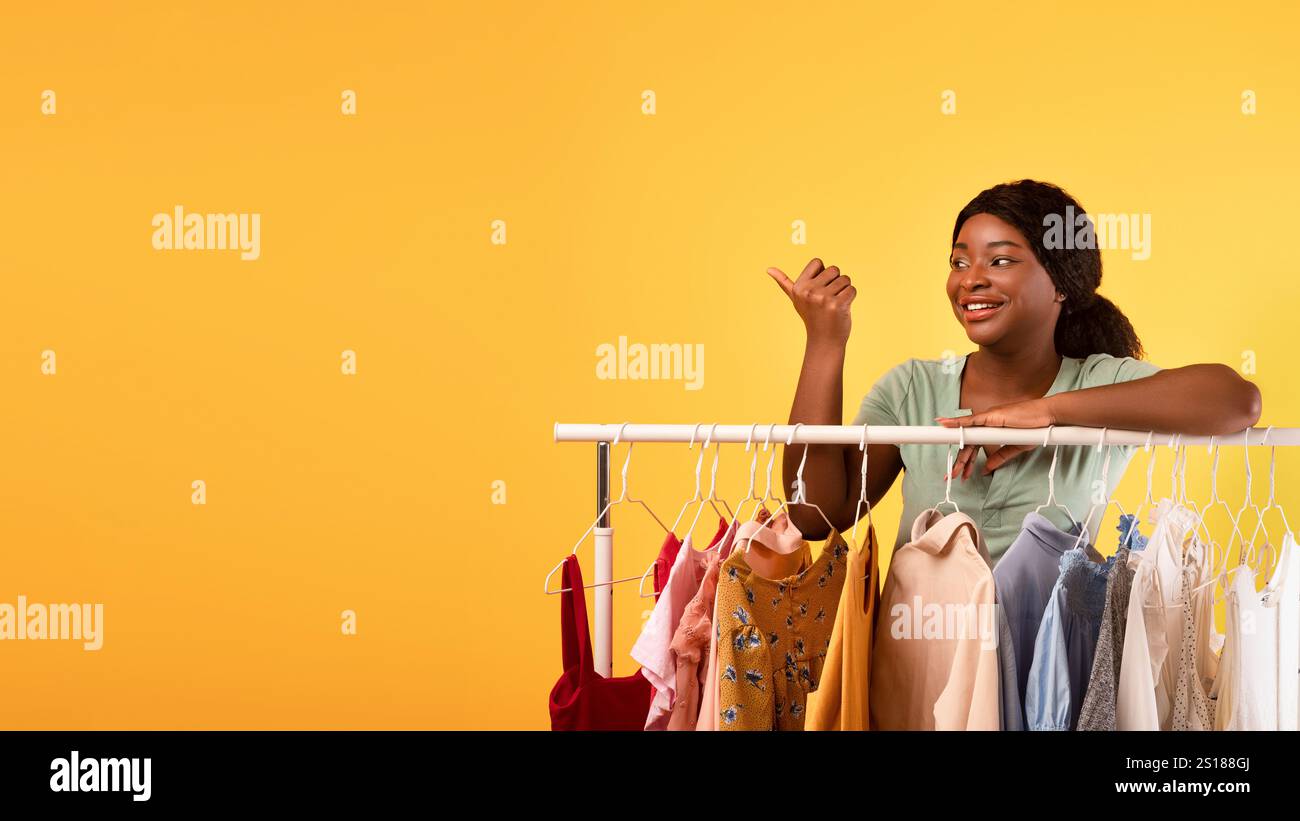 Portrait of young black woman standing near clothing rack, pointing at ...