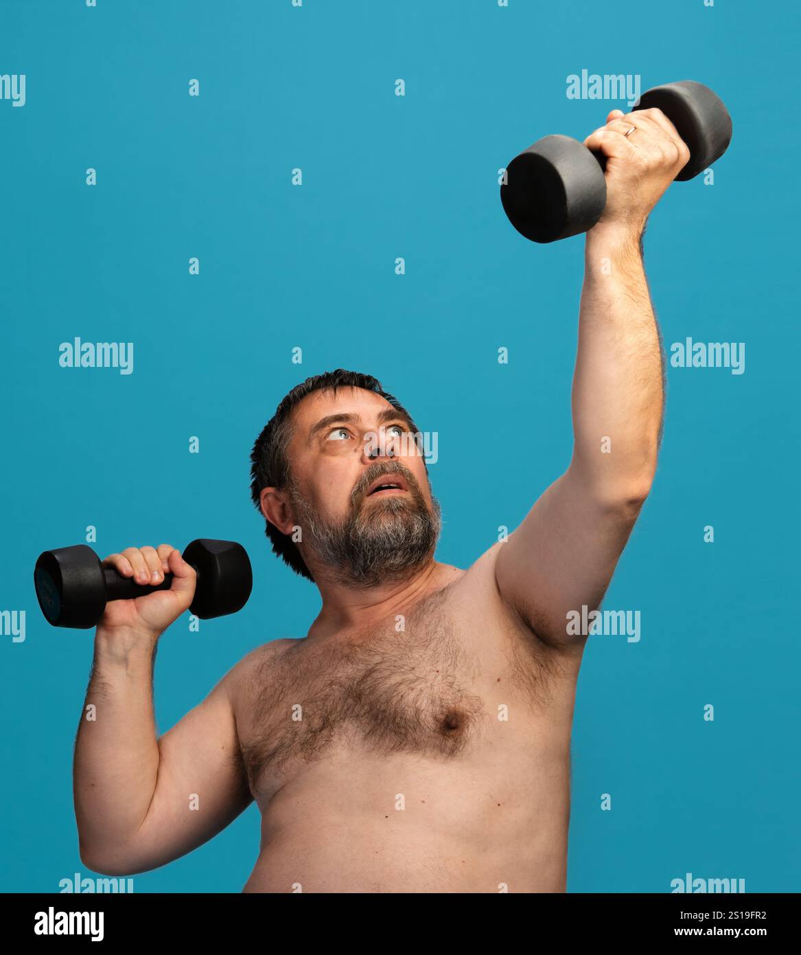 An elderly man without a shirt lifts dumbbells while exercising. His focused expression conveys ...