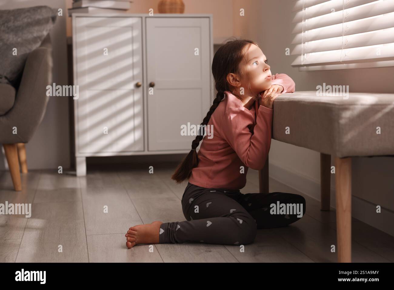 Orphanage concept. Sad girl and looking through window blinds indoors ...