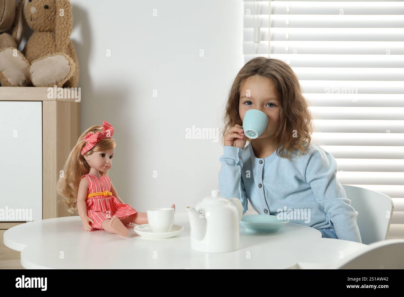 Cute little girl playing tea party with doll at white table in room ...