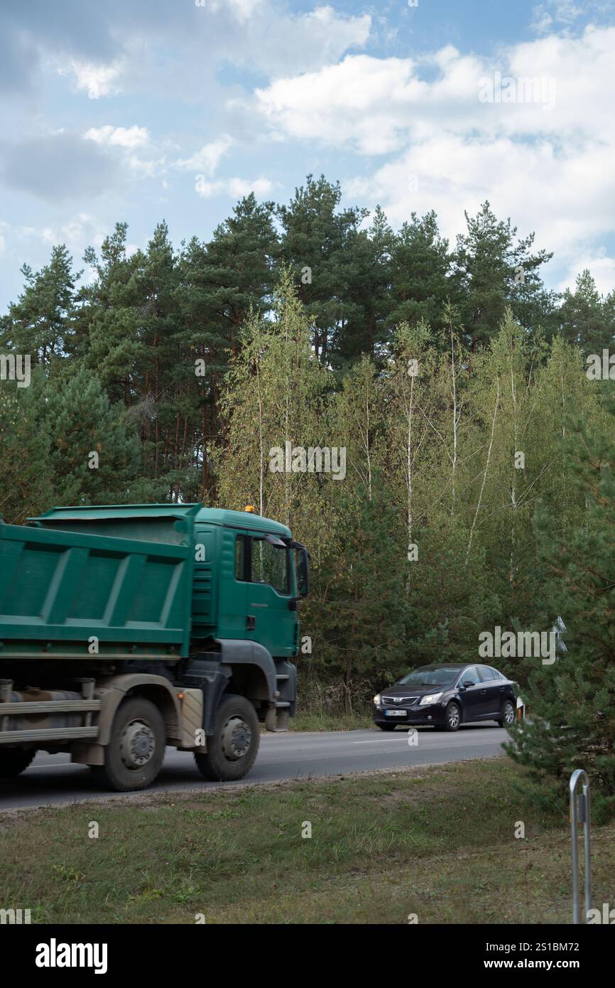 The image shows a green dump truck on a road, with a black car nearby ...