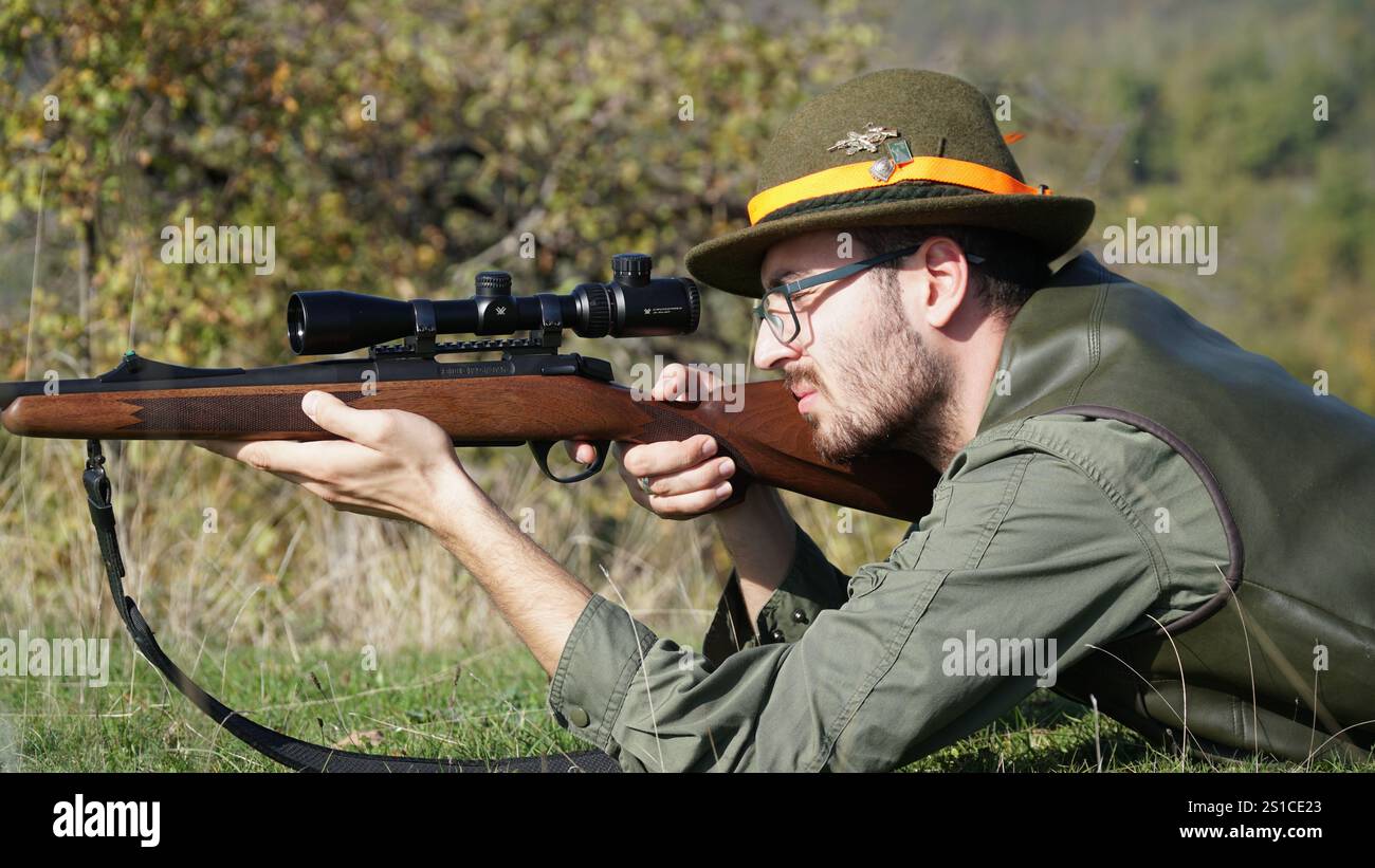 Hunter in camouflage aims his rifle in a dense forest, blending with ...