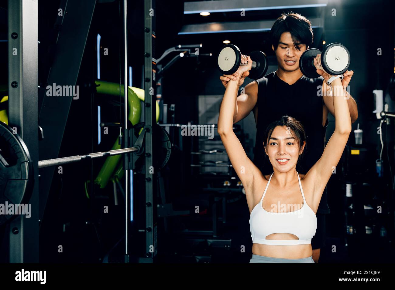 Fit Asian woman lifting dumbbells with a smile, supported by her ...
