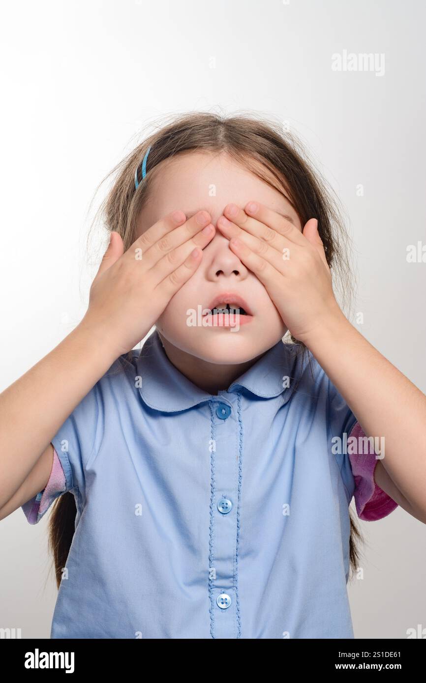 A cute little girl in a light blue shirt playfully covers her eyes with ...
